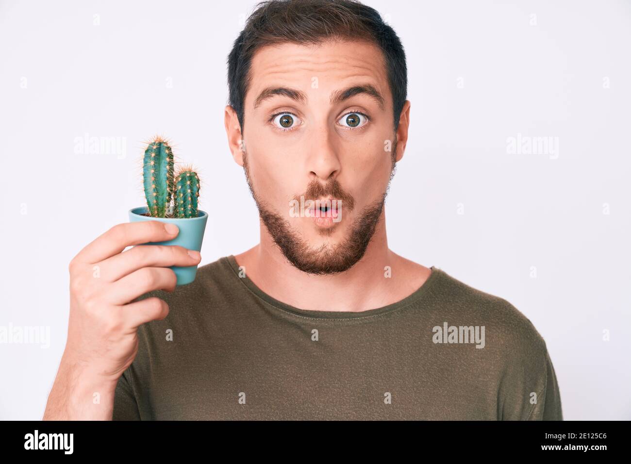 Young handsome man holding small cactus pot scared and amazed with open ...