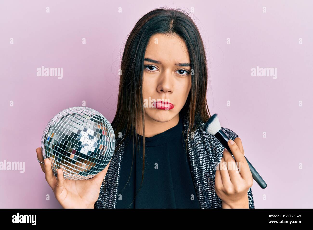Young hispanic girl holding shiny disco ball and makeup brush depressed ...