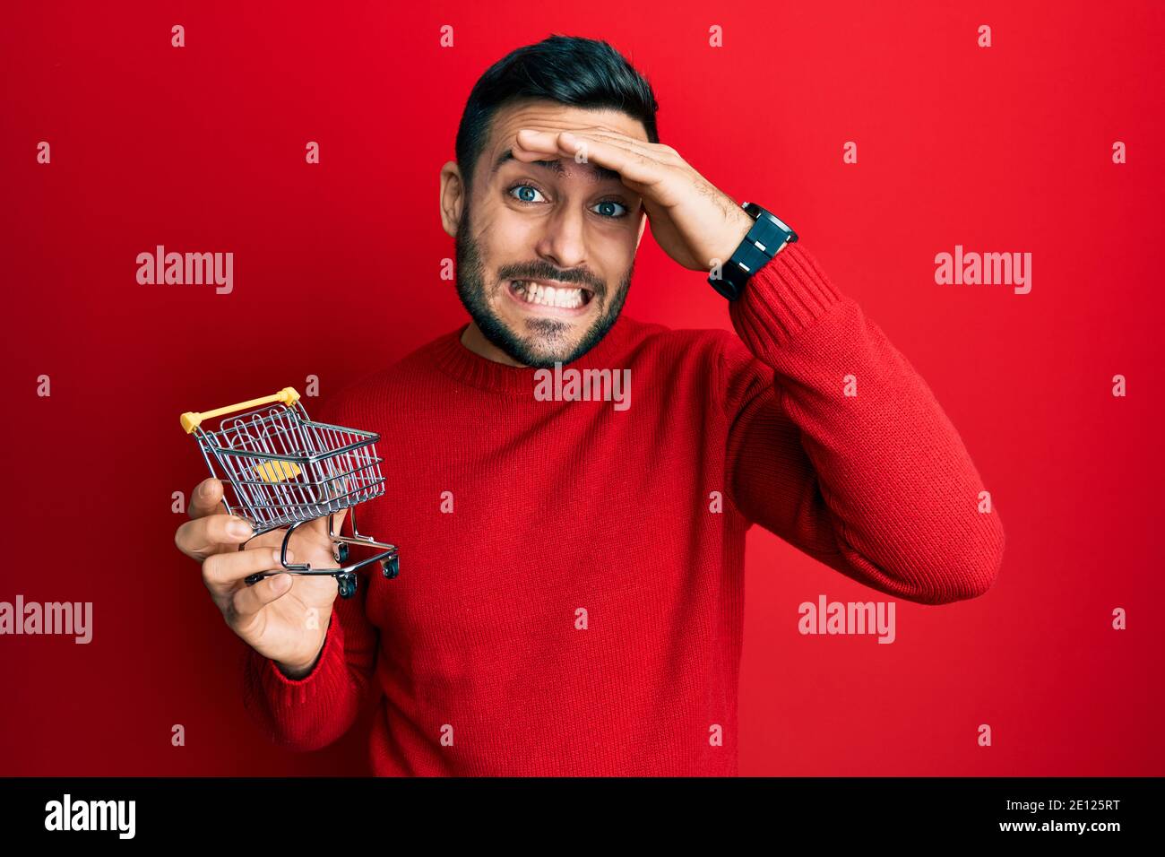 Young hispanic man holding small supermarket shopping cart stressed and ...