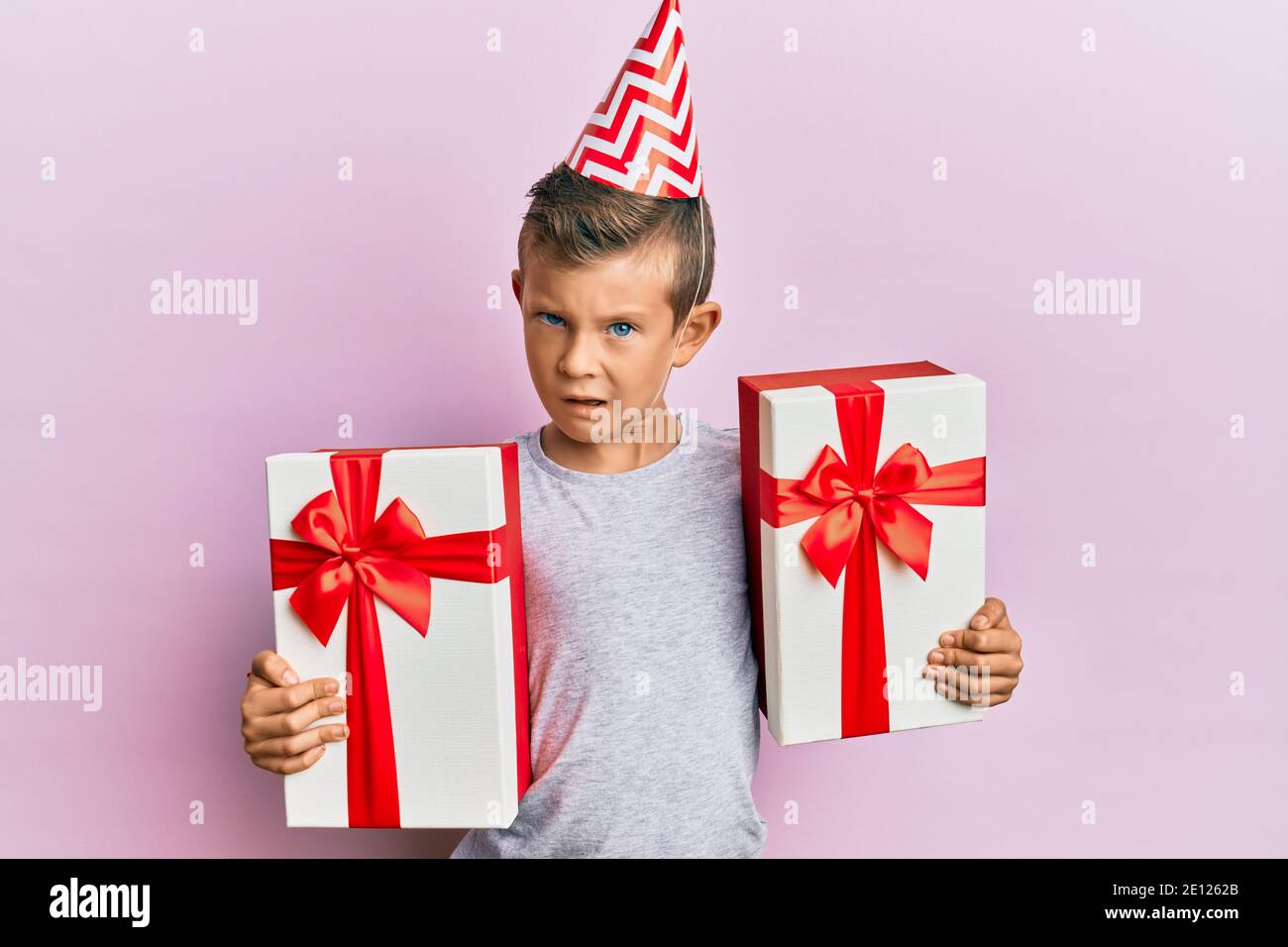 Adorable caucasian kid wearing birthday hat holding presents in shock ...