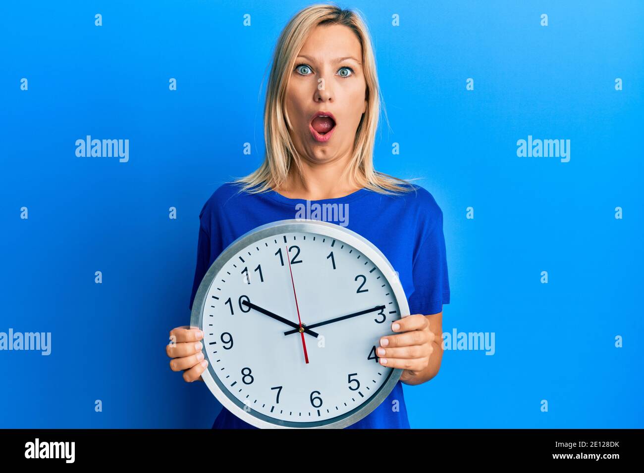 Beautiful middle age blonde woman holding big clock afraid and shocked ...