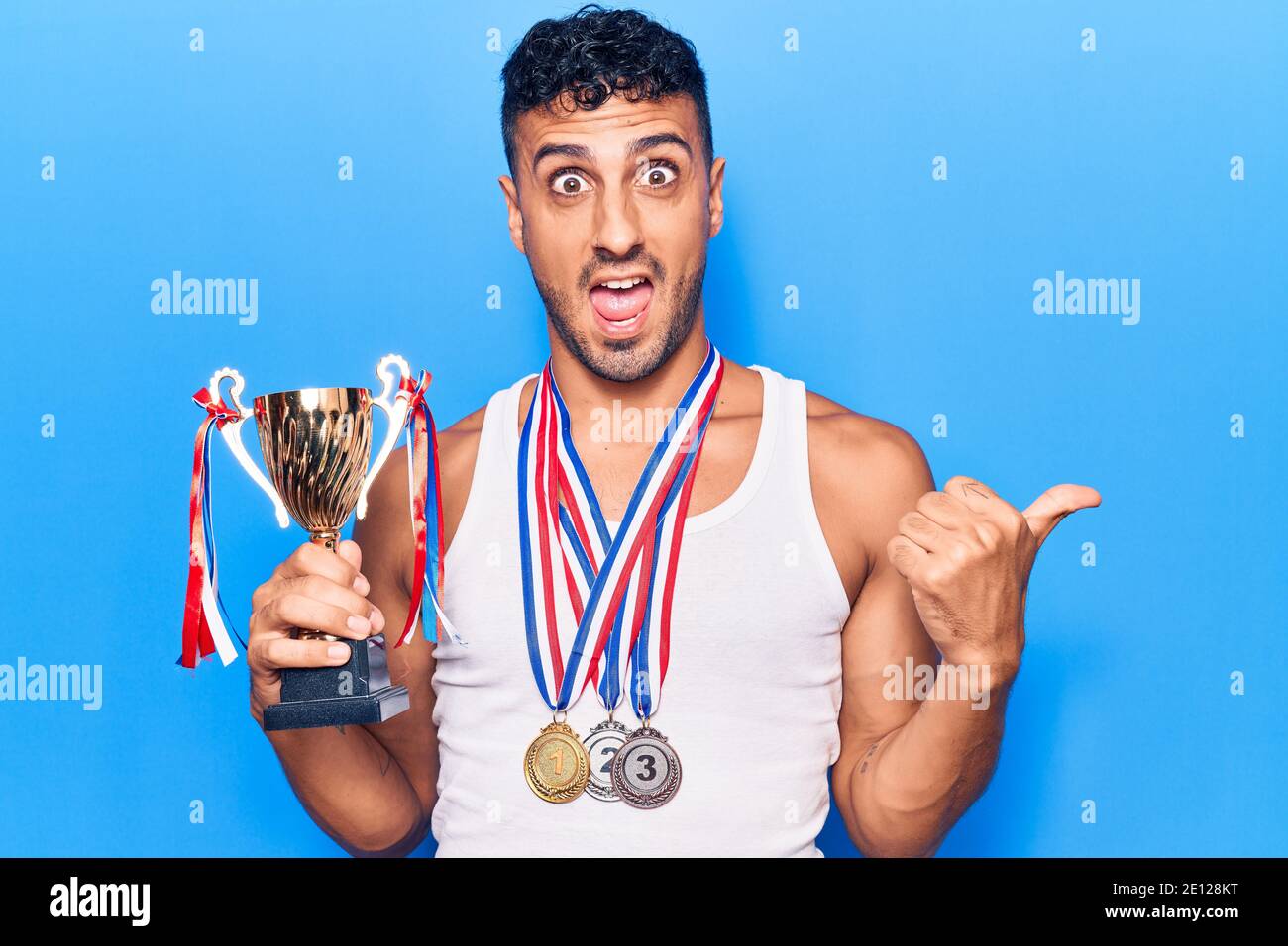 Young hispanic man wearing winner medals holding trophy pointing thumb ...