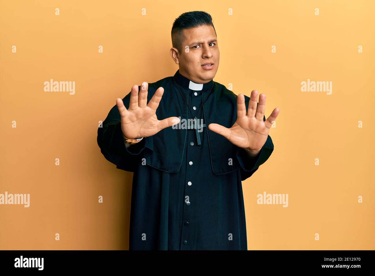 Young latin priest man standing over yellow background afraid and ...