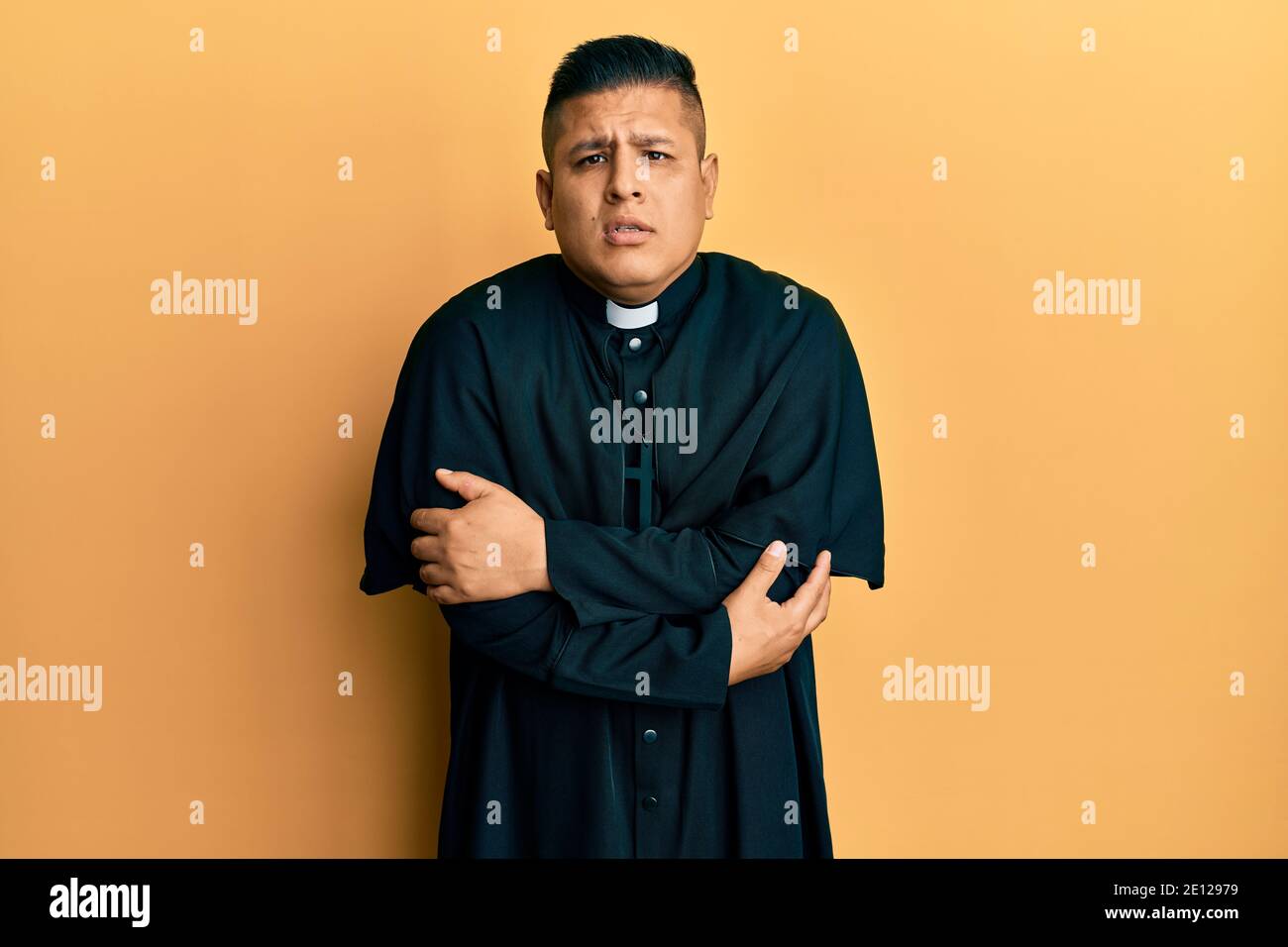 Young latin priest man standing over yellow background shaking and ...