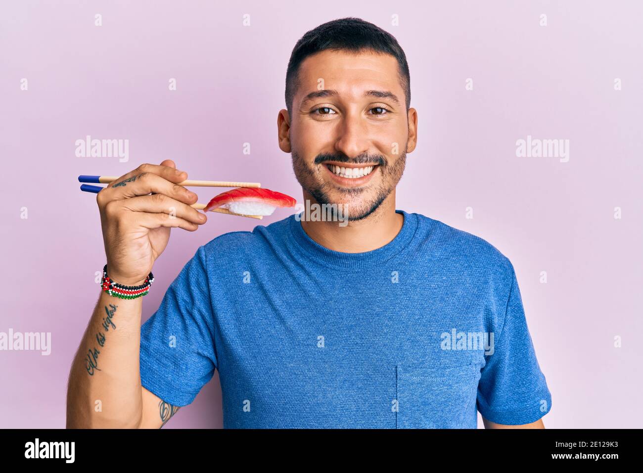 Handsome man with tattoos eating tuna sushi using chopsticks looking ...