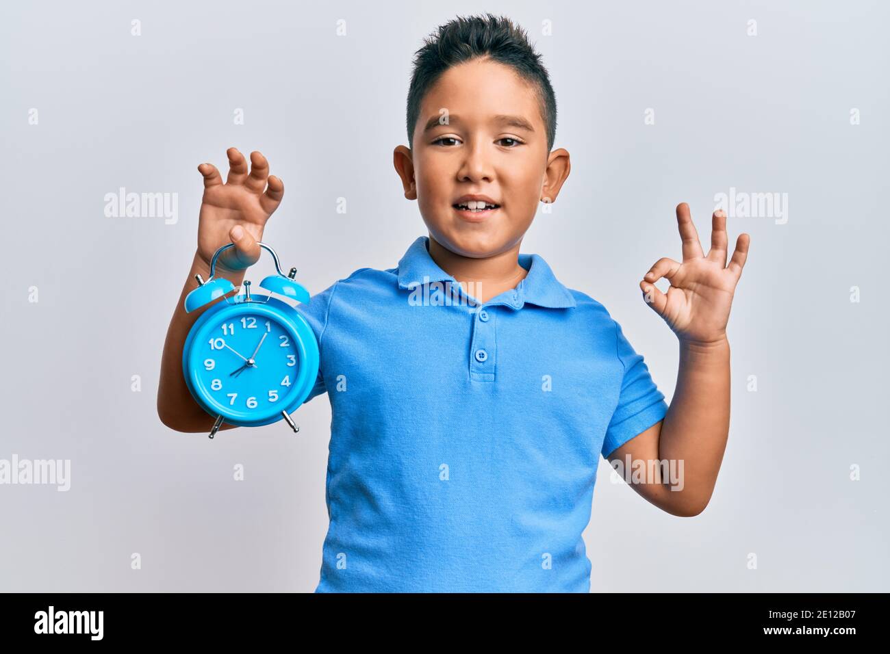 Little boy hispanic kid holding alarm clock doing ok sign with fingers ...