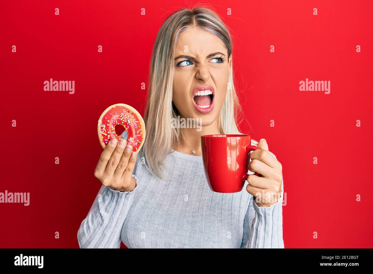 Beautiful blonde woman eating doughnut and drinking coffee angry and ...