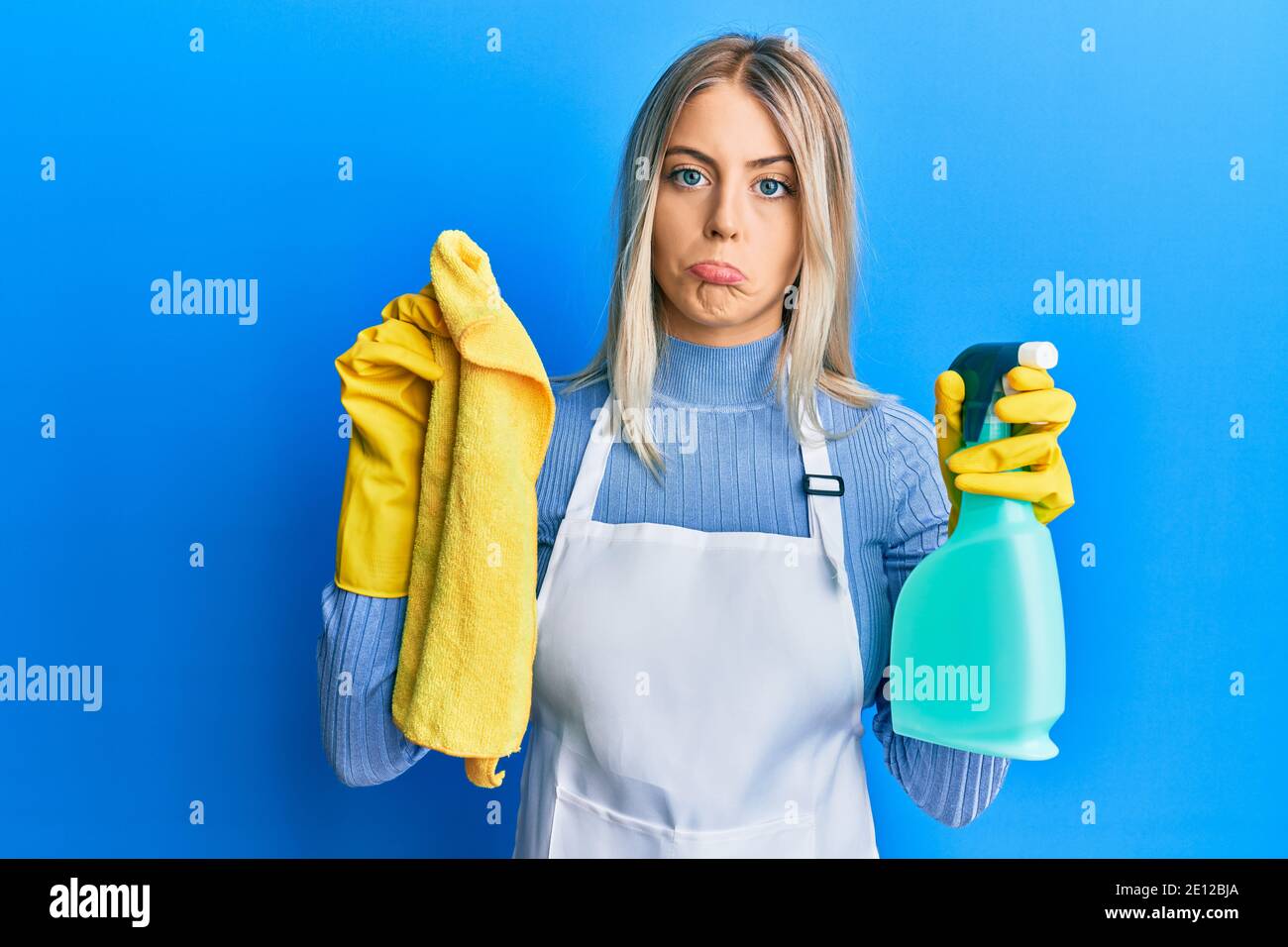 Beautiful blonde woman wearing cleaner apron holding cleaning products ...