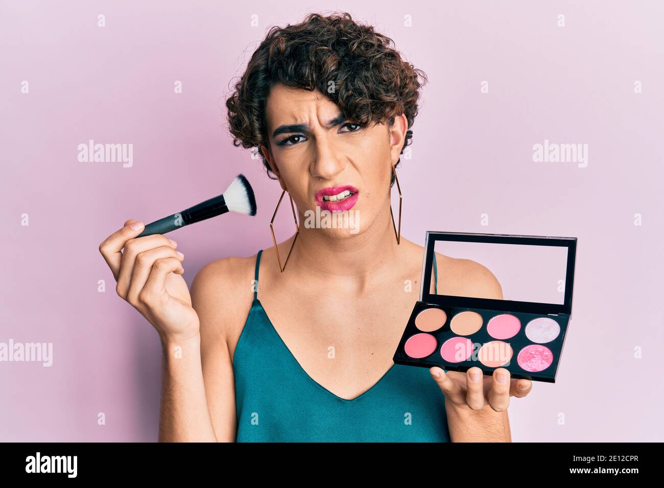 Young man wearing woman make up holding makeup brush and blush in shock ...