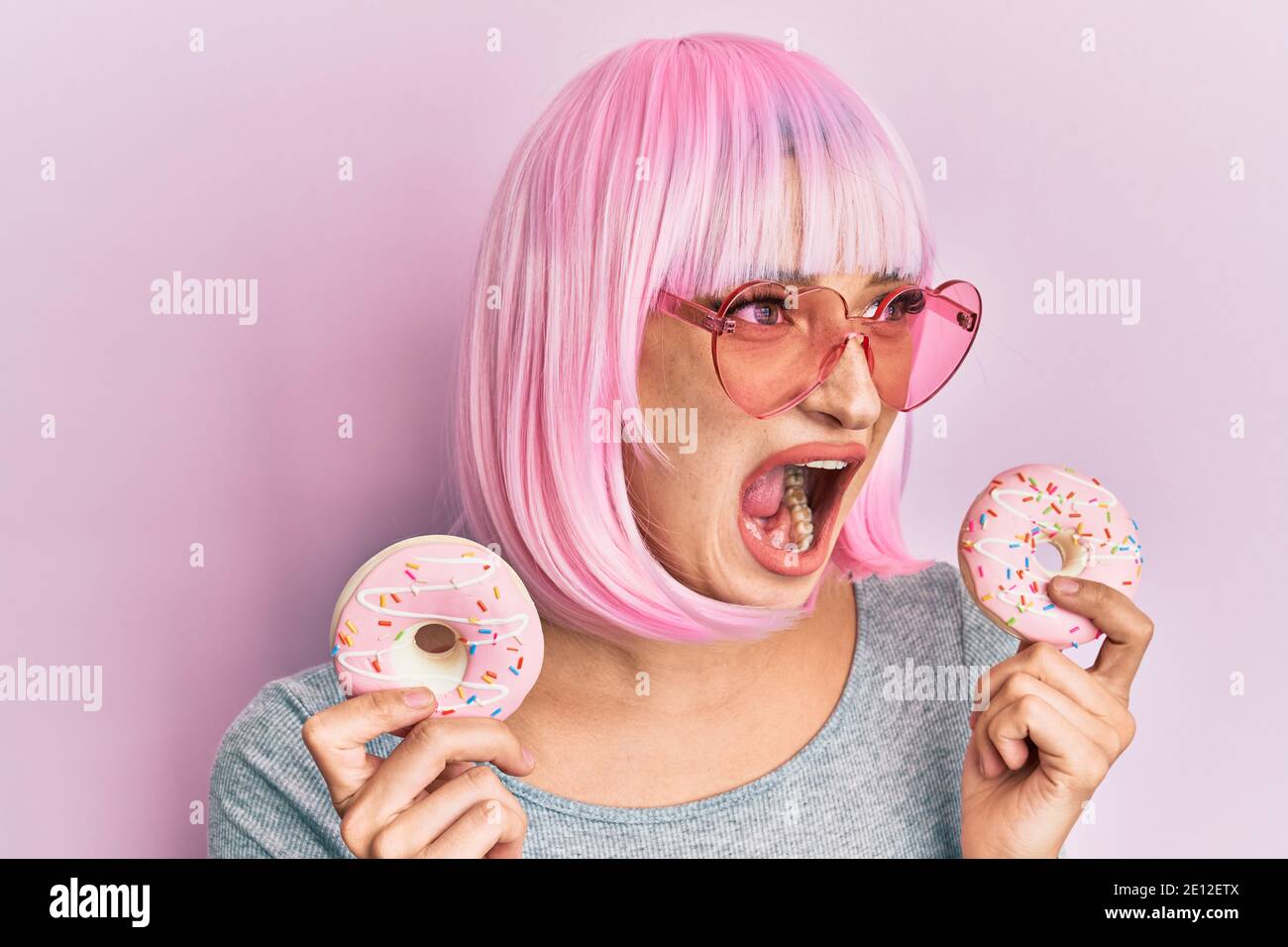 Young caucasian woman wearing pink wig holding donuts angry and mad ...