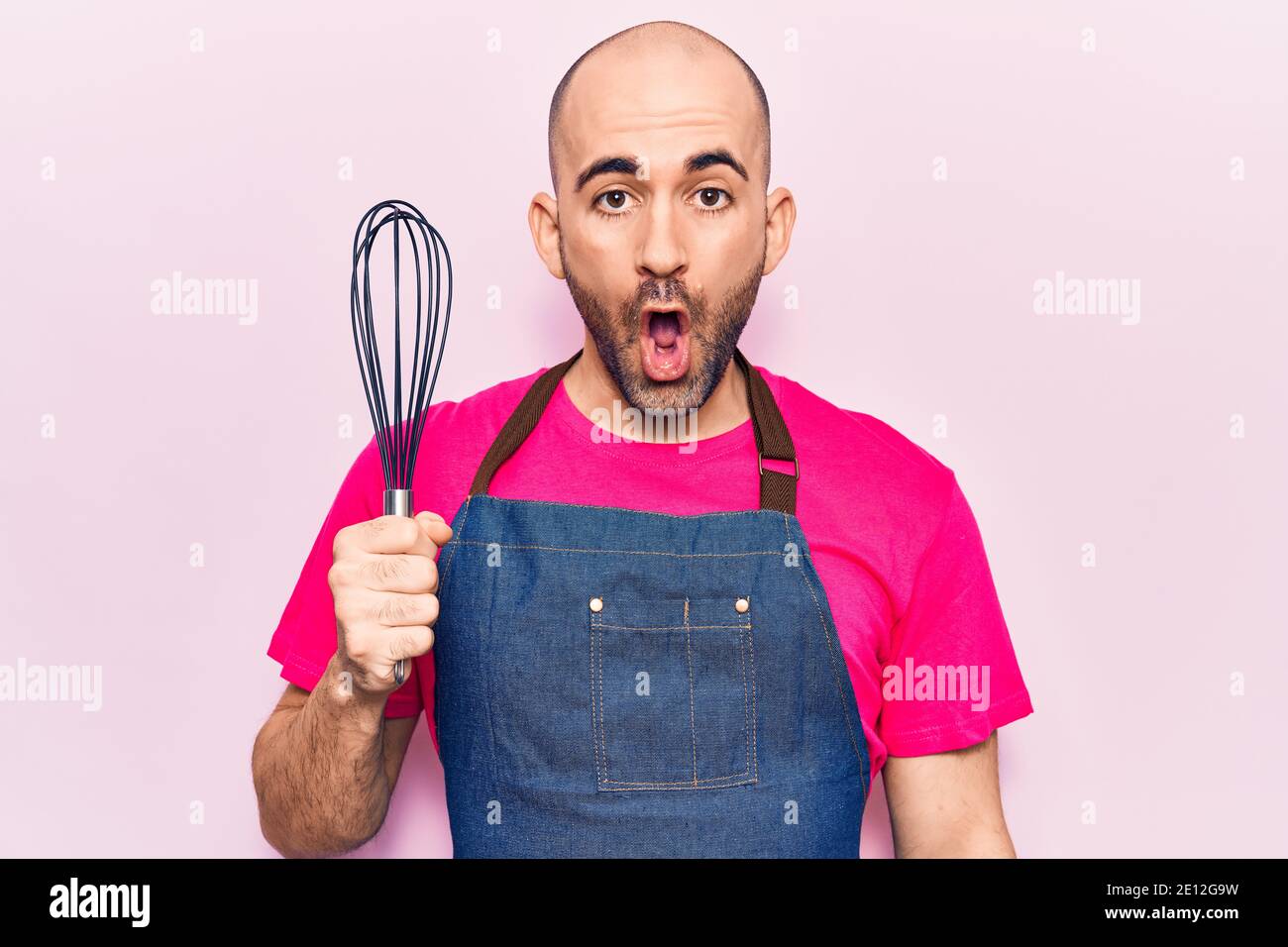 Young handsome bald man wearing apron holding whisk scared and amazed ...