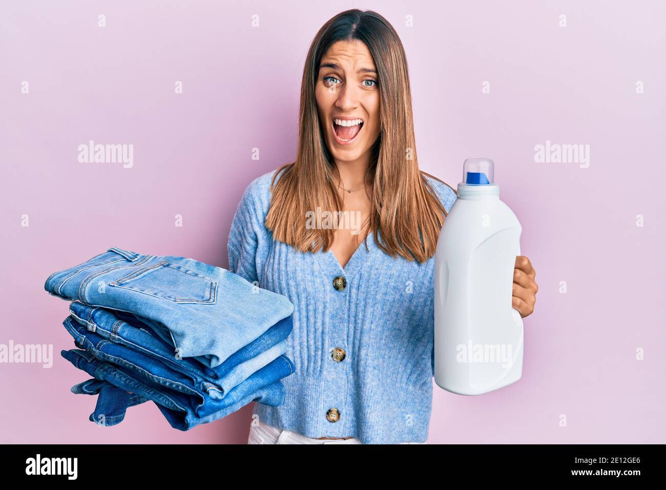 Brunette young woman holding jeans for laundry and detergent bottle celebrating crazy and amazed ...