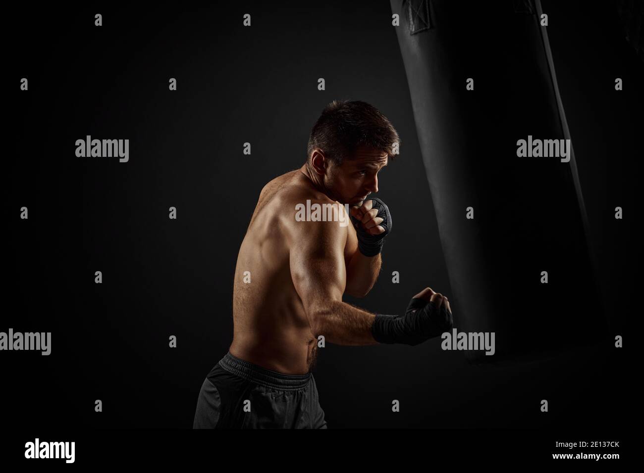 Aggressive shirtless boxer punching in boxing bag on black background ...