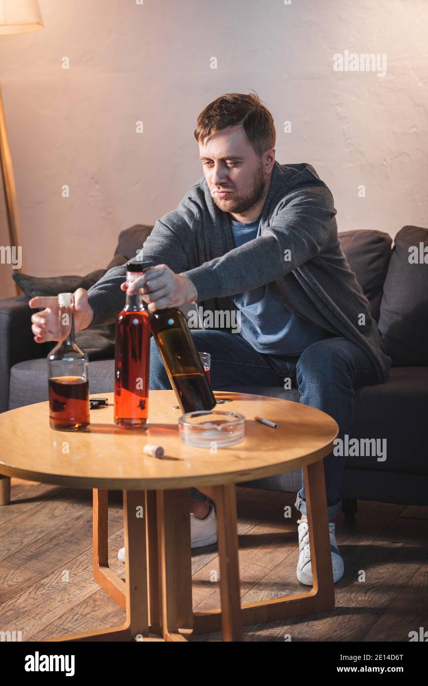 drunk man taking bottles of alcohol from table while sitting at home ...