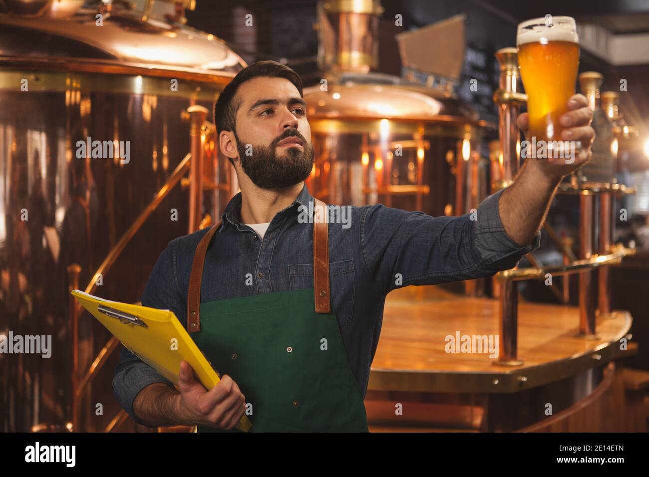 Bearded professional brewer holding clipboard, examining freshly made ...