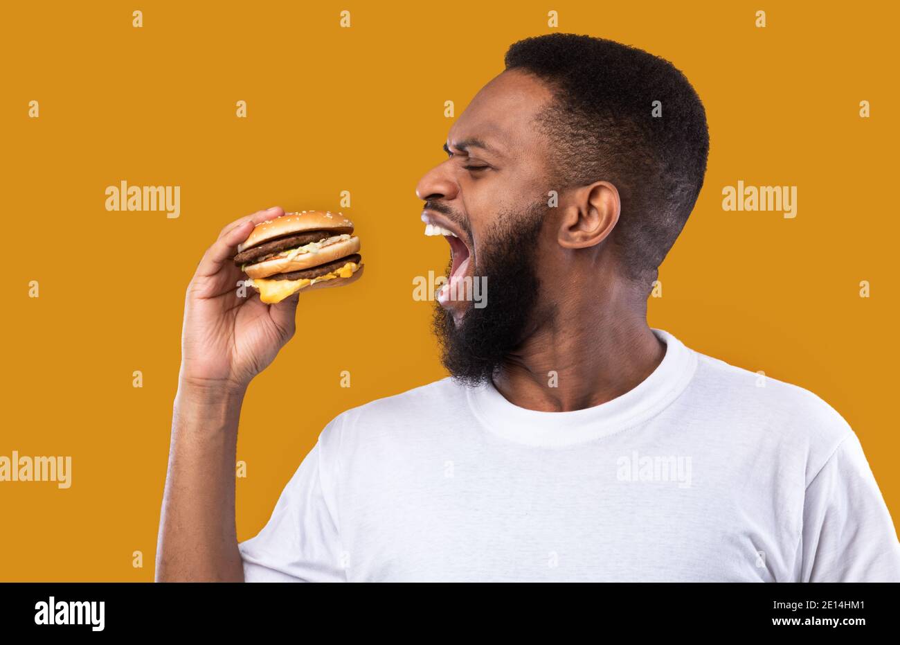 Hungry African Guy Biting Burger Posing On Yellow Background, Side-View ...