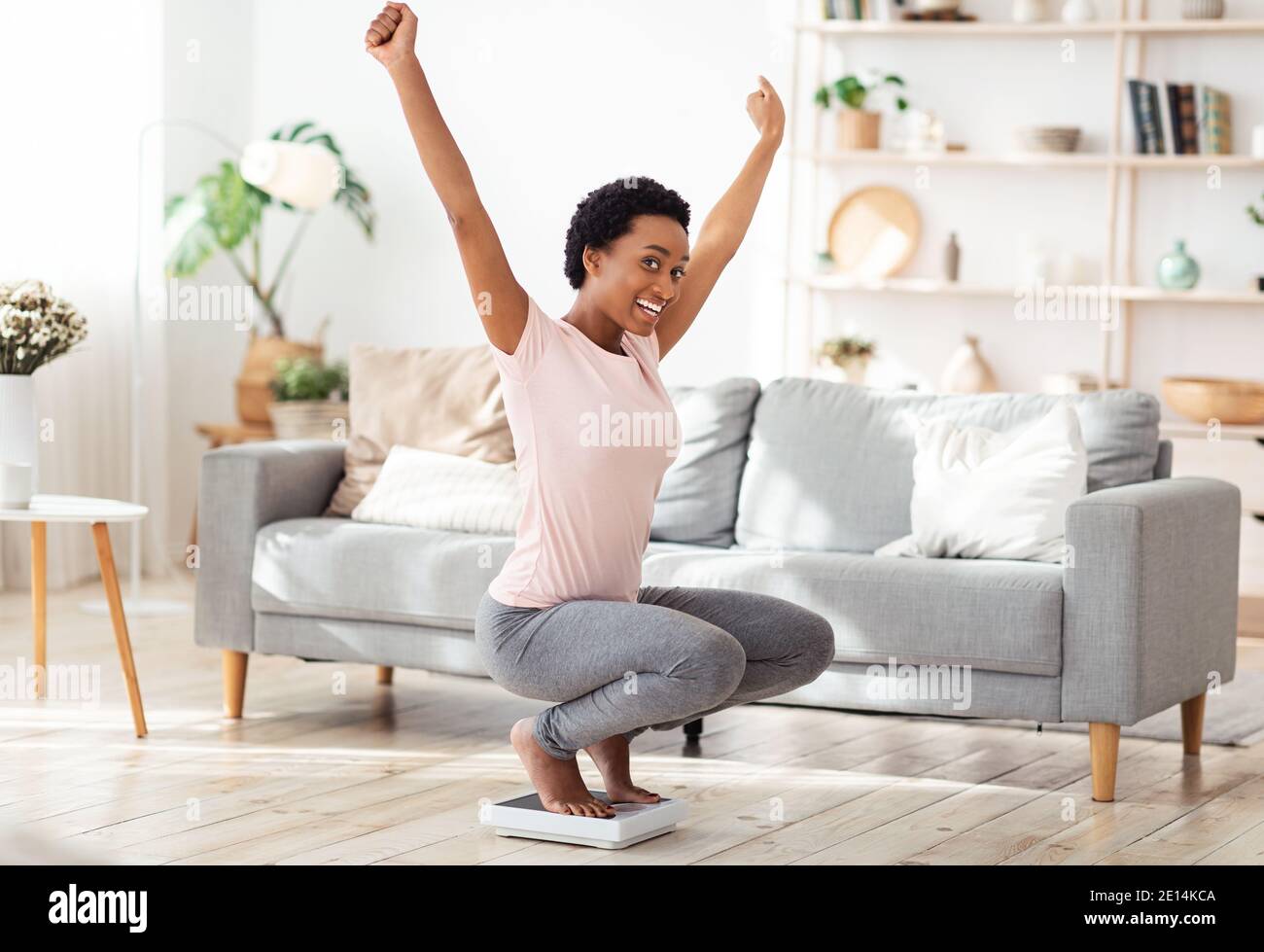 Young black woman sitting on scales, raising hands up in excitement ...