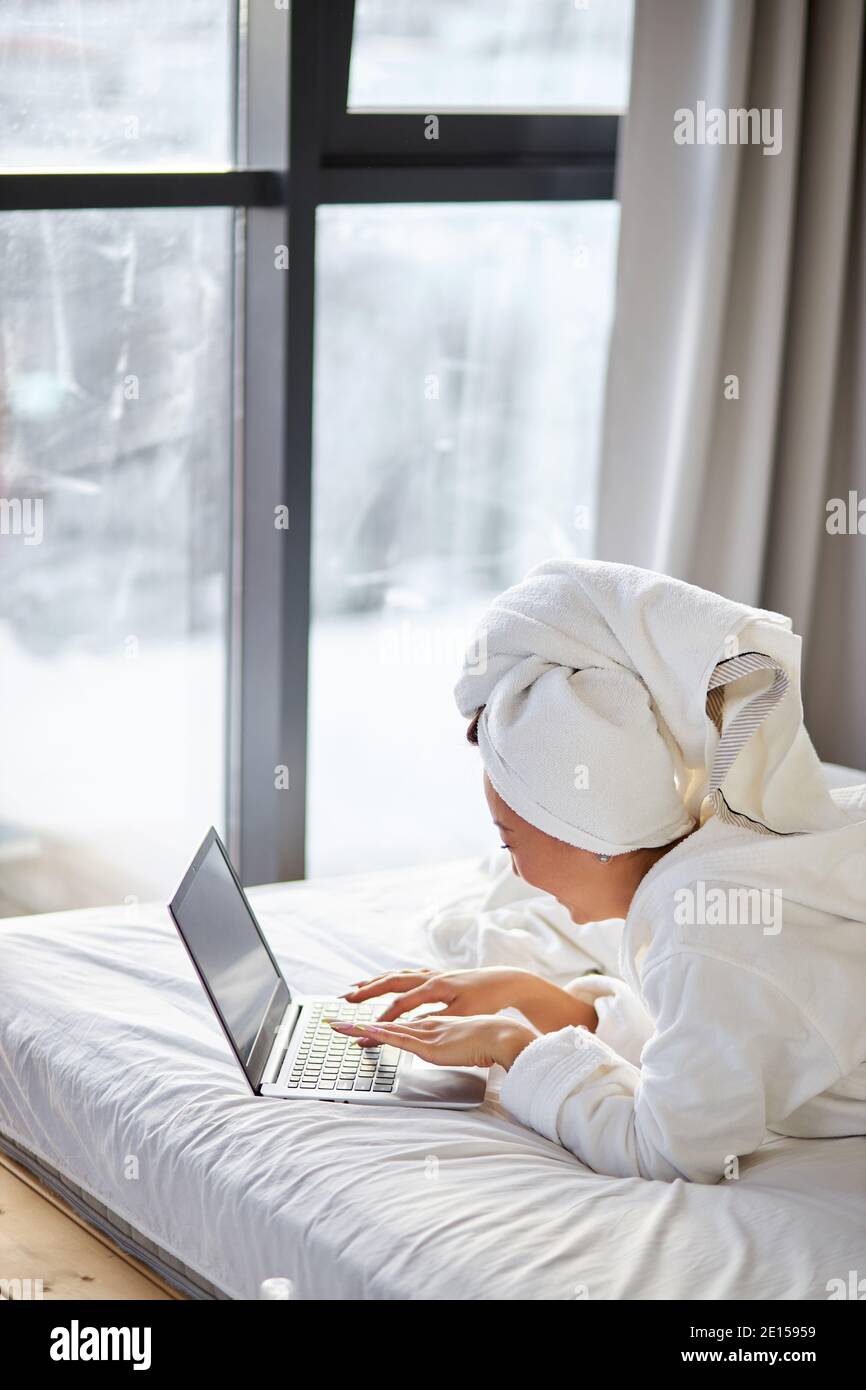 woman in white bathrobe in bedroom at home typing message to friends ...