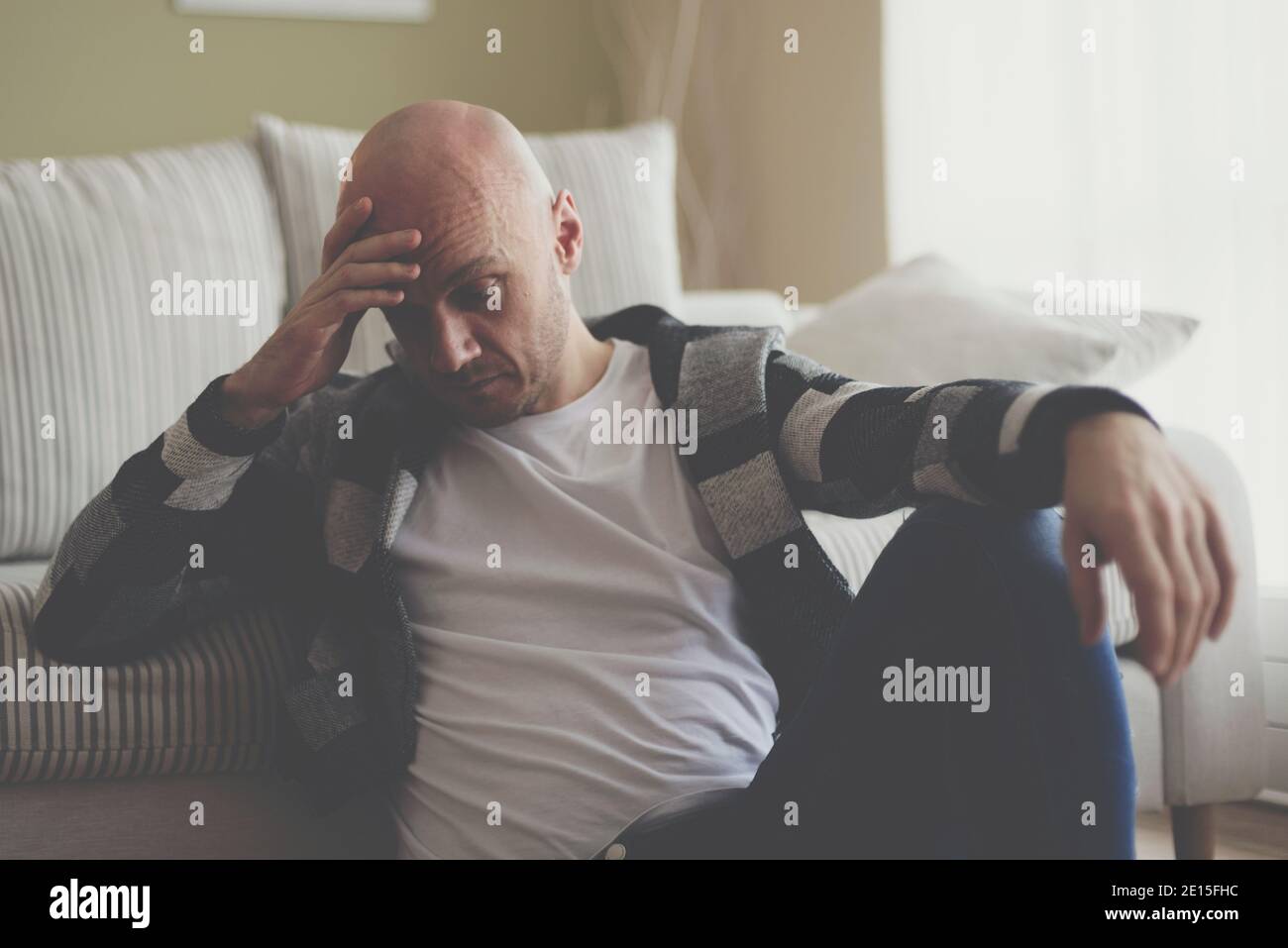A depressed man sits on the living room floor covering his face with ...