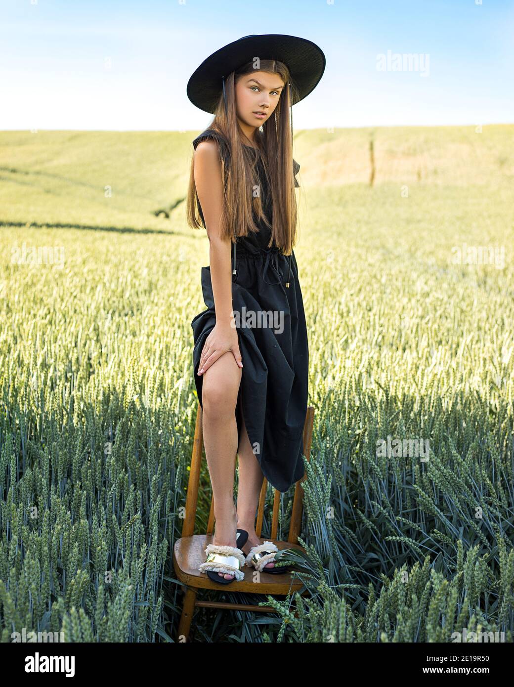 Beautiful girl in field standing on chair in dress and black hat ...
