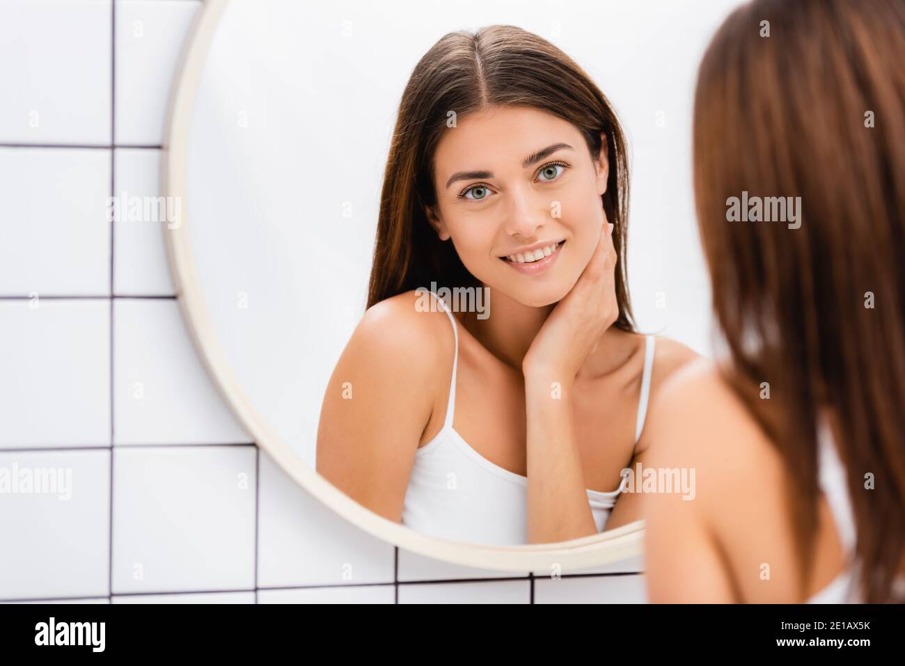 Happy Young Woman Touching Face And Smiling Near Mirror In Bathroom