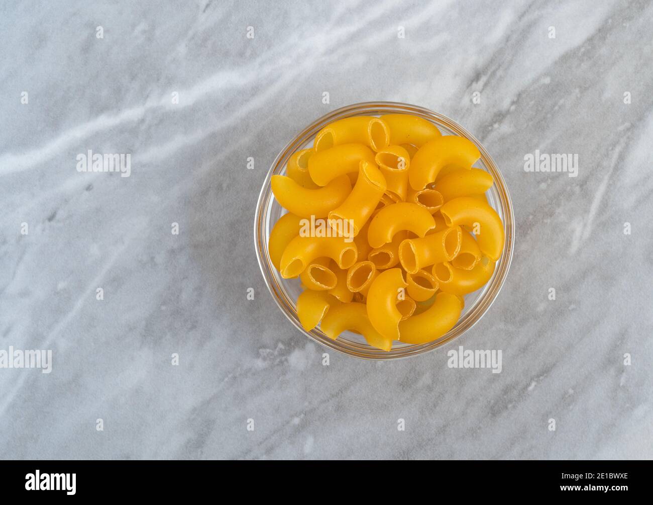 Organic elbow macaroni in a glass bowl on a gray countertop illuminated ...