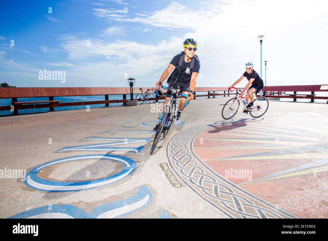 Two male cyclists ride bicycles on a platform above Lake Ontario on one ...