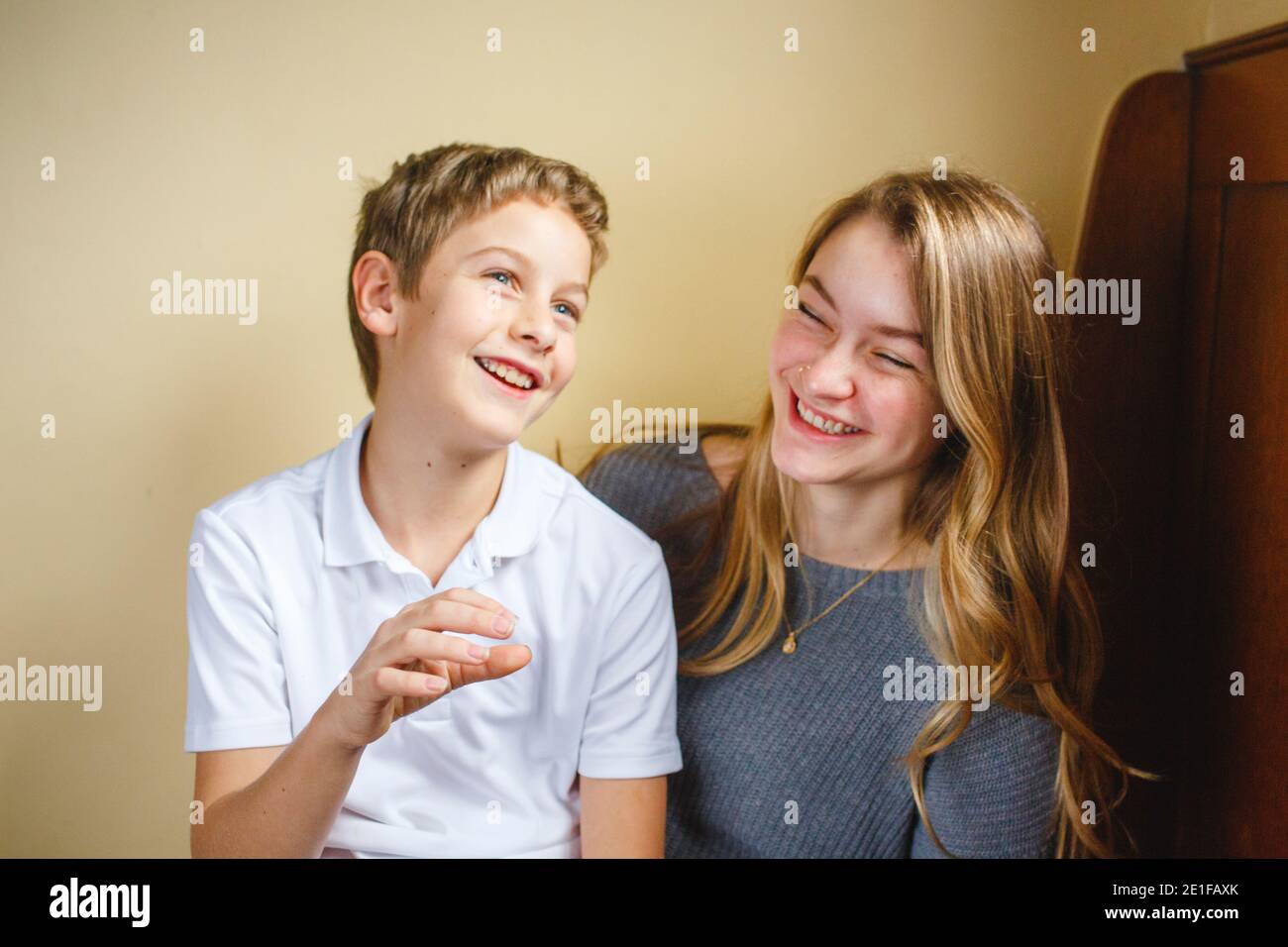 Two siblings sit together on a wood bench laughing with joy indoors ...