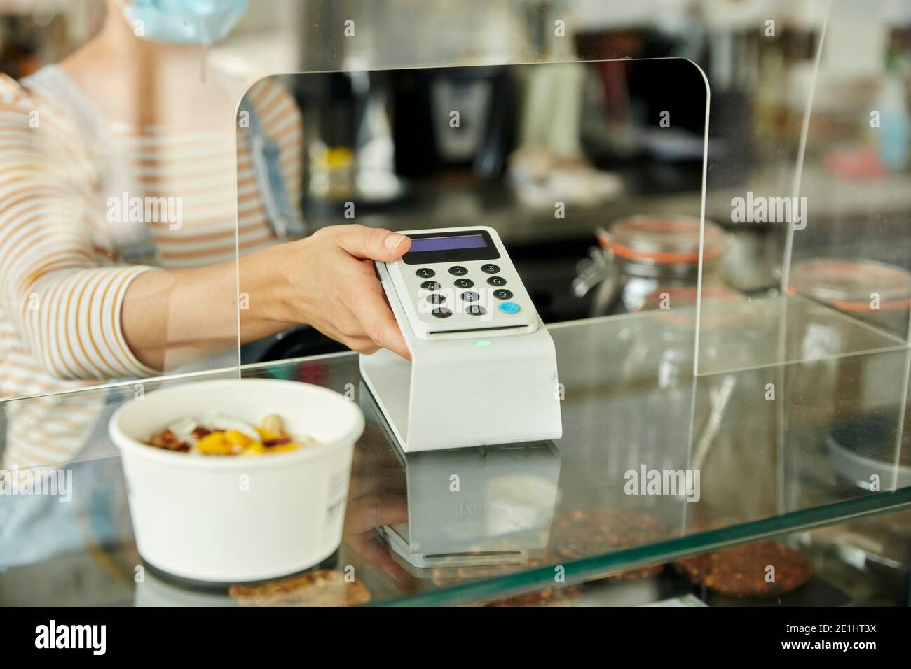 Woman in a face mask behind cafe counter with safety screen, offering a ...