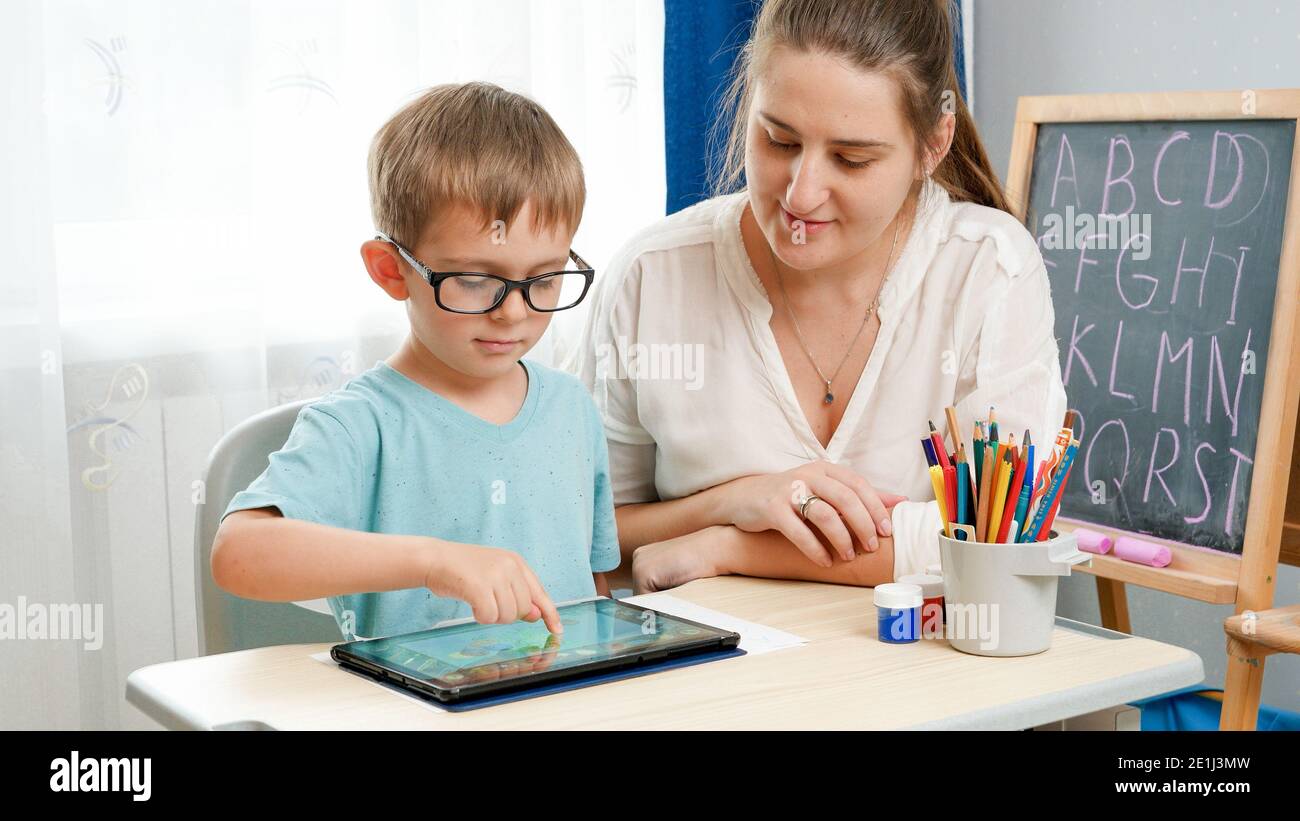 Little boy in eyeglasses playing on tablet computer. Mother hugging her ...