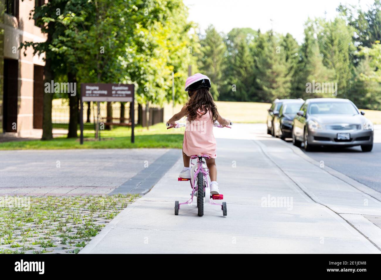 Young girl in pink dress and helmet riding pink bike on the street ...