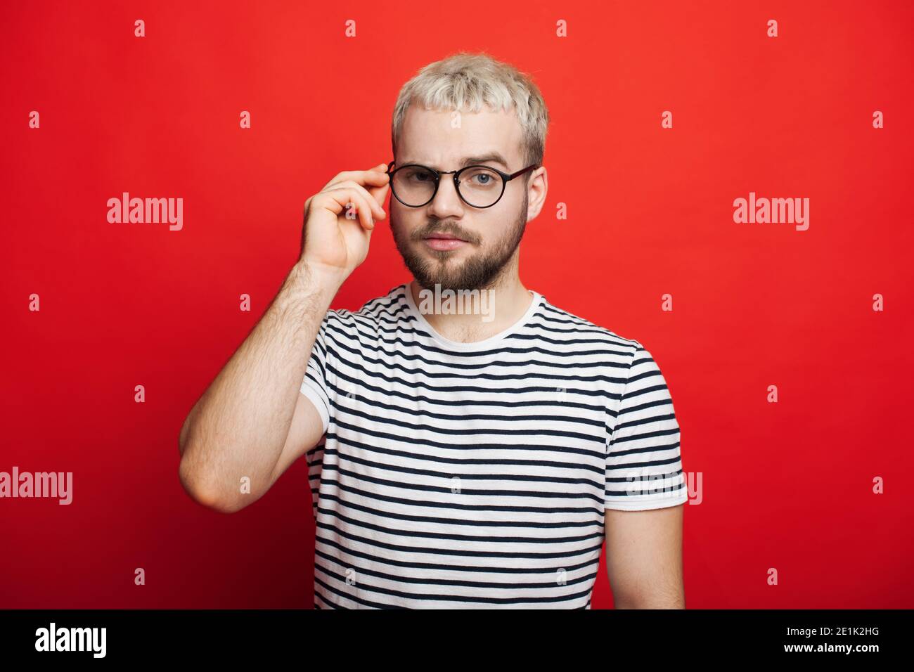 Serious man is touching his glasses posing on a red studio wall looking ...