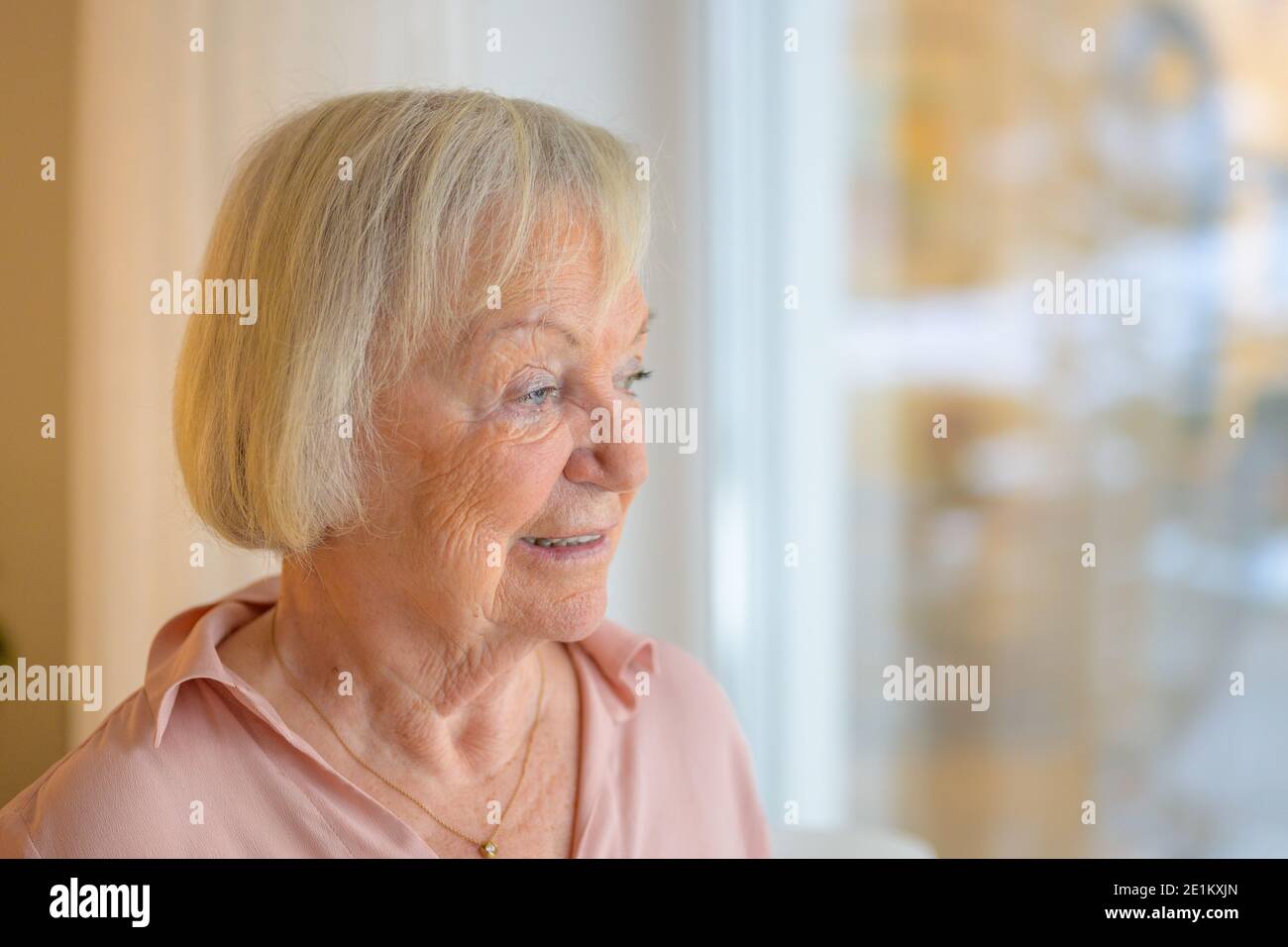 Thoughtful senior lady staring out of a window watching outside in a ...