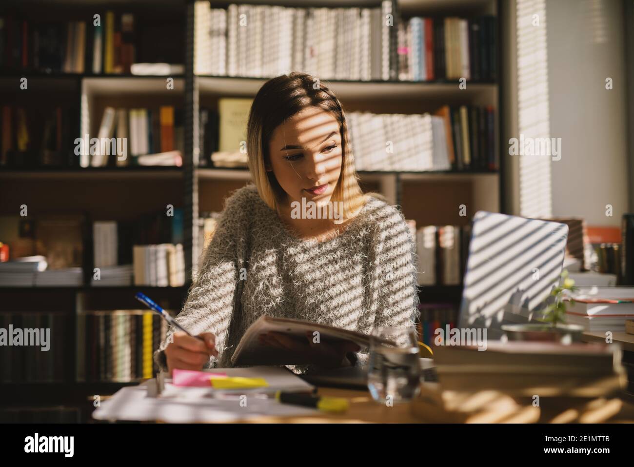 Cute young school girl sitting in library and taking notes while ...