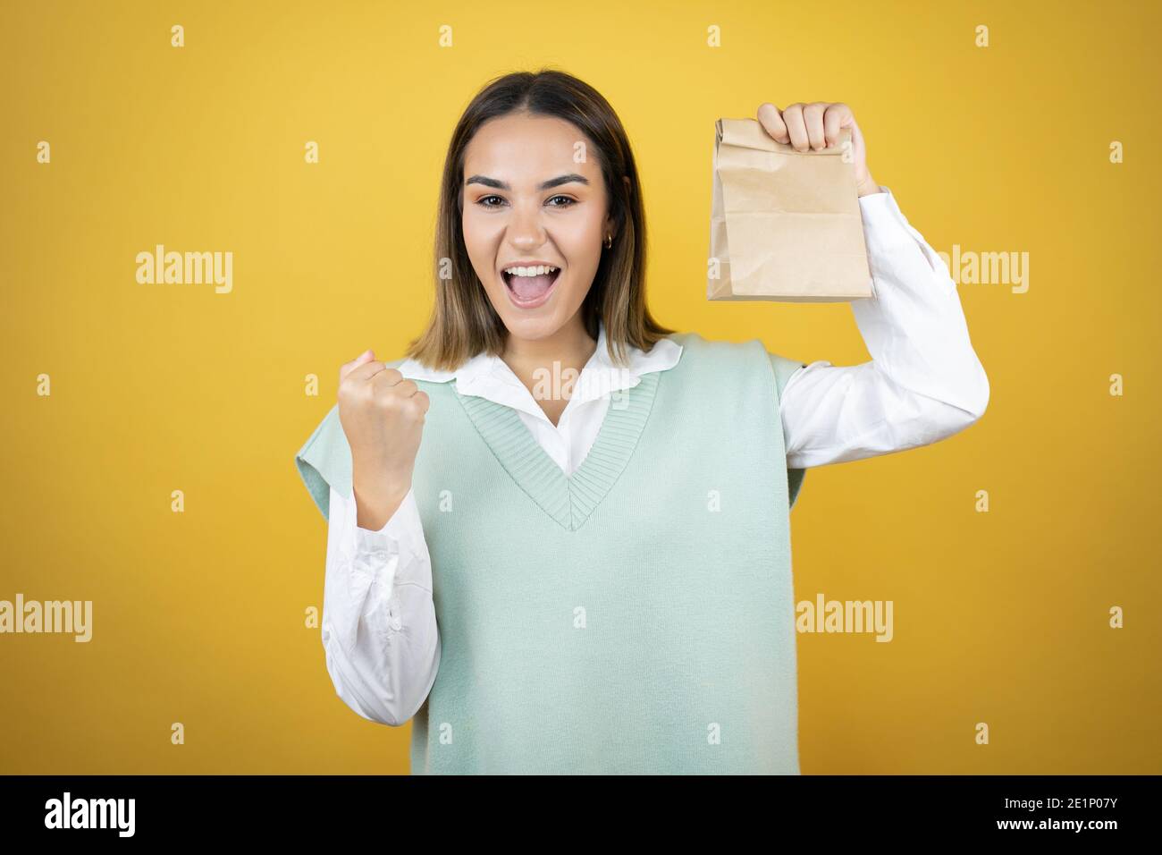 Pretty young woman standing over yellow background holding a paper bag ...