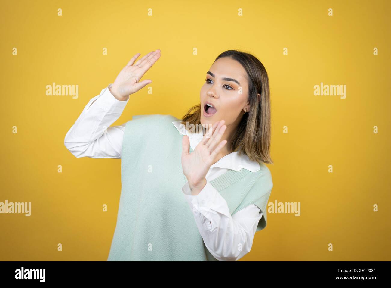 Pretty young woman standing over yellow background scared with her arms ...