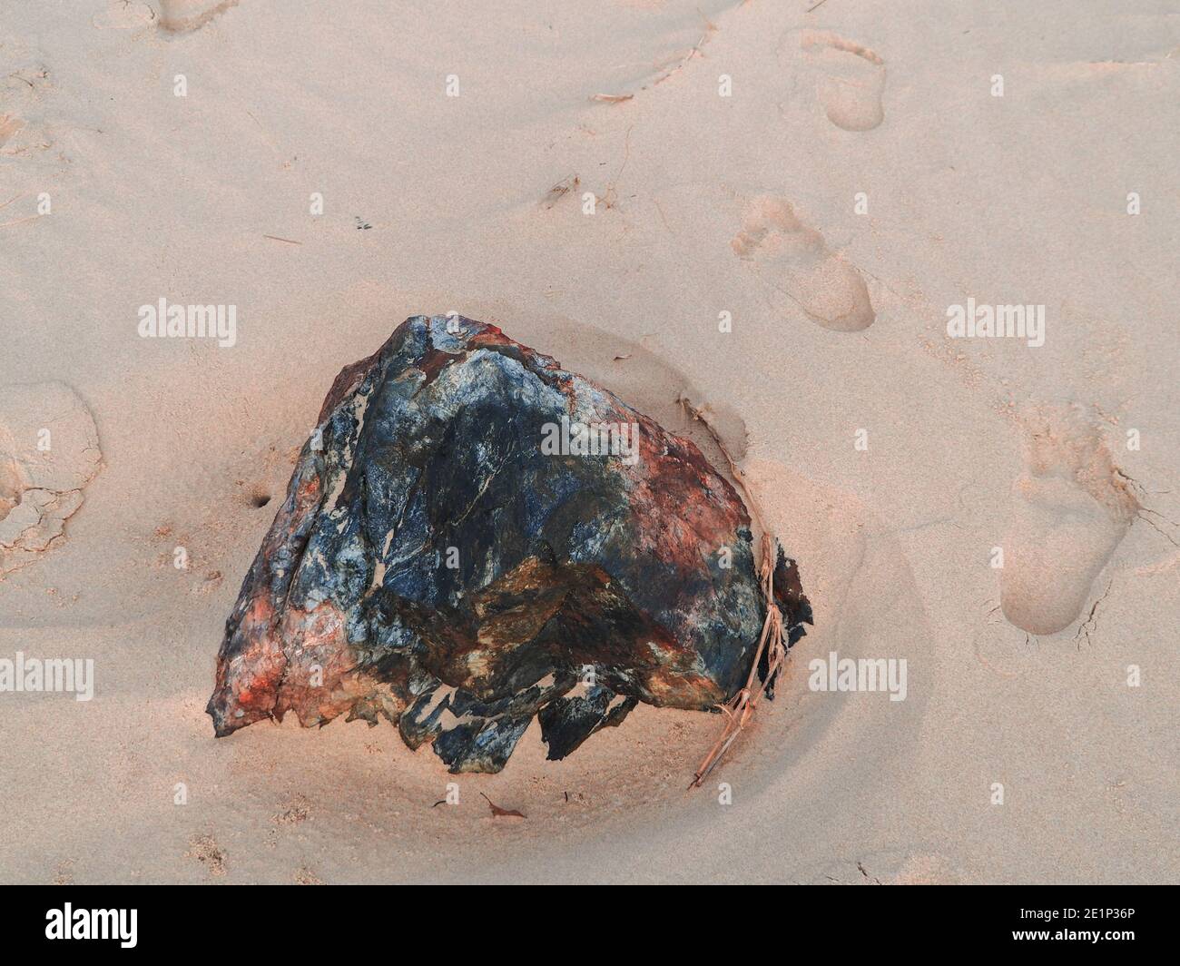 A dark grey and pinkish red rock on the sandy beach surrounded by human ...