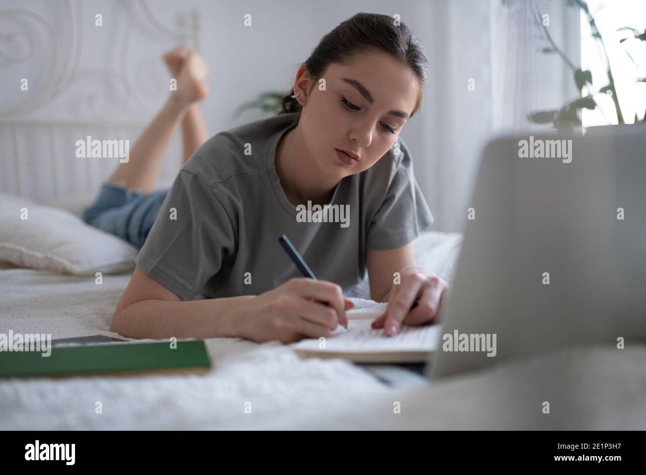 Serious female student lying and writing in notebook while working on ...