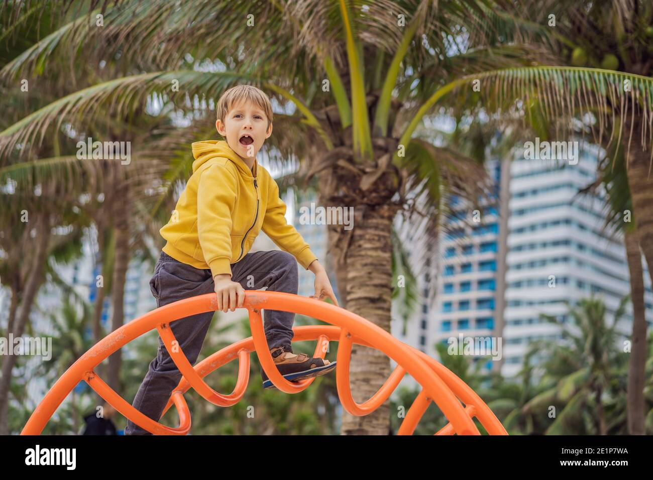The boy climbs on the playground against the background of palm trees ...