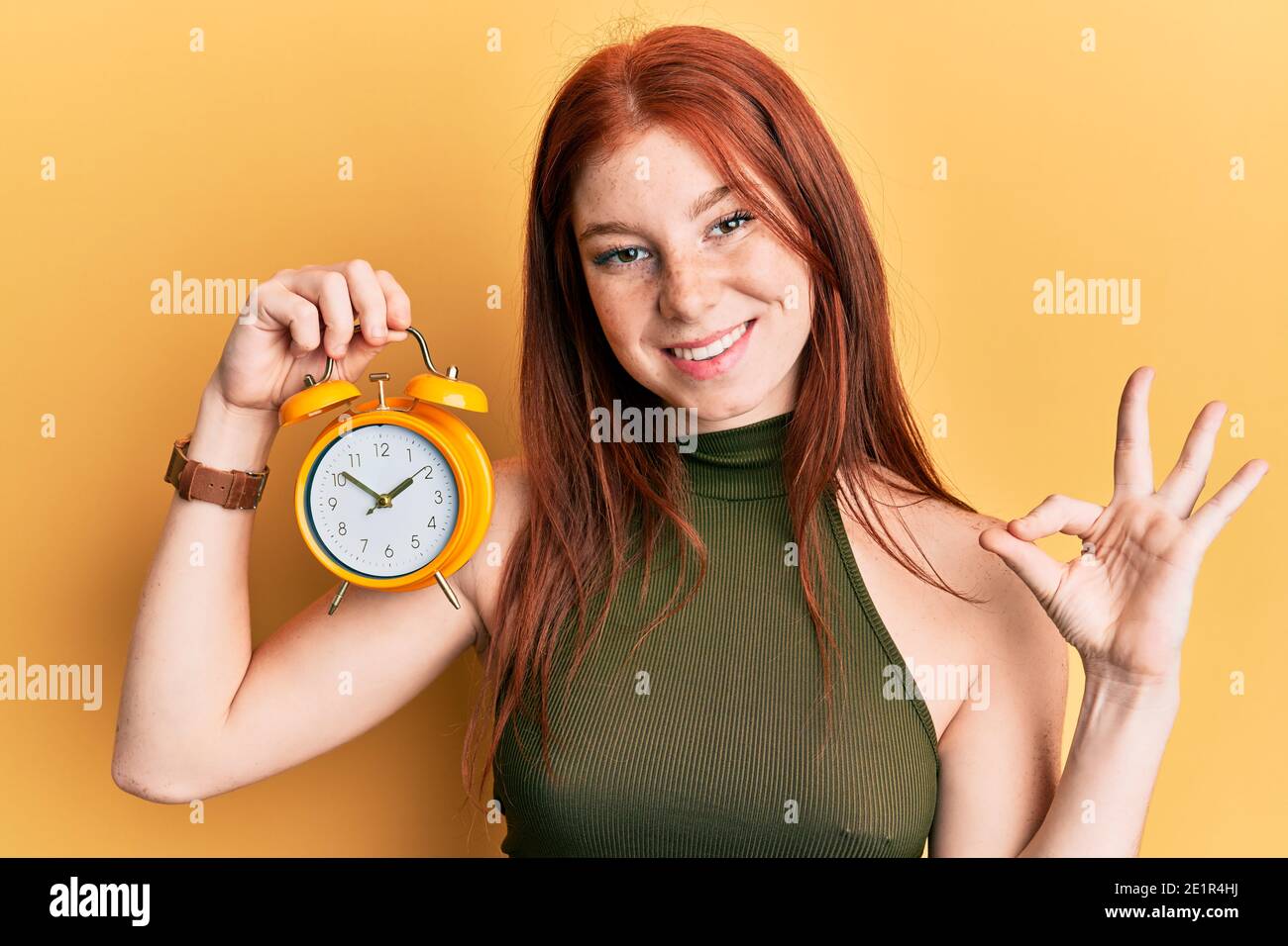 Young red head girl holding alarm clock doing ok sign with fingers ...
