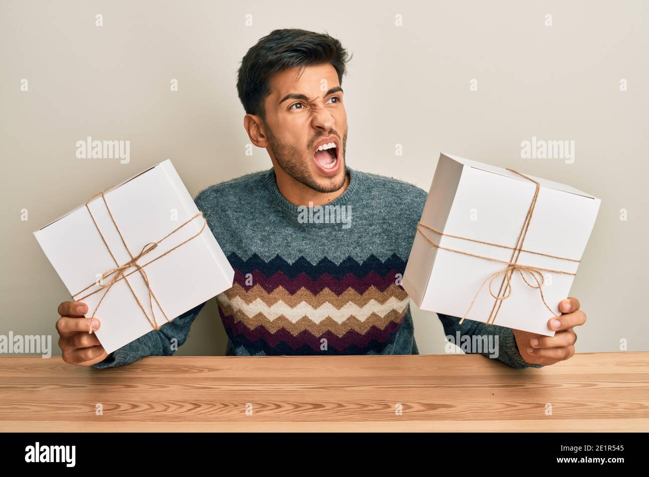 Young handsome man holding gifts angry and mad screaming frustrated and ...
