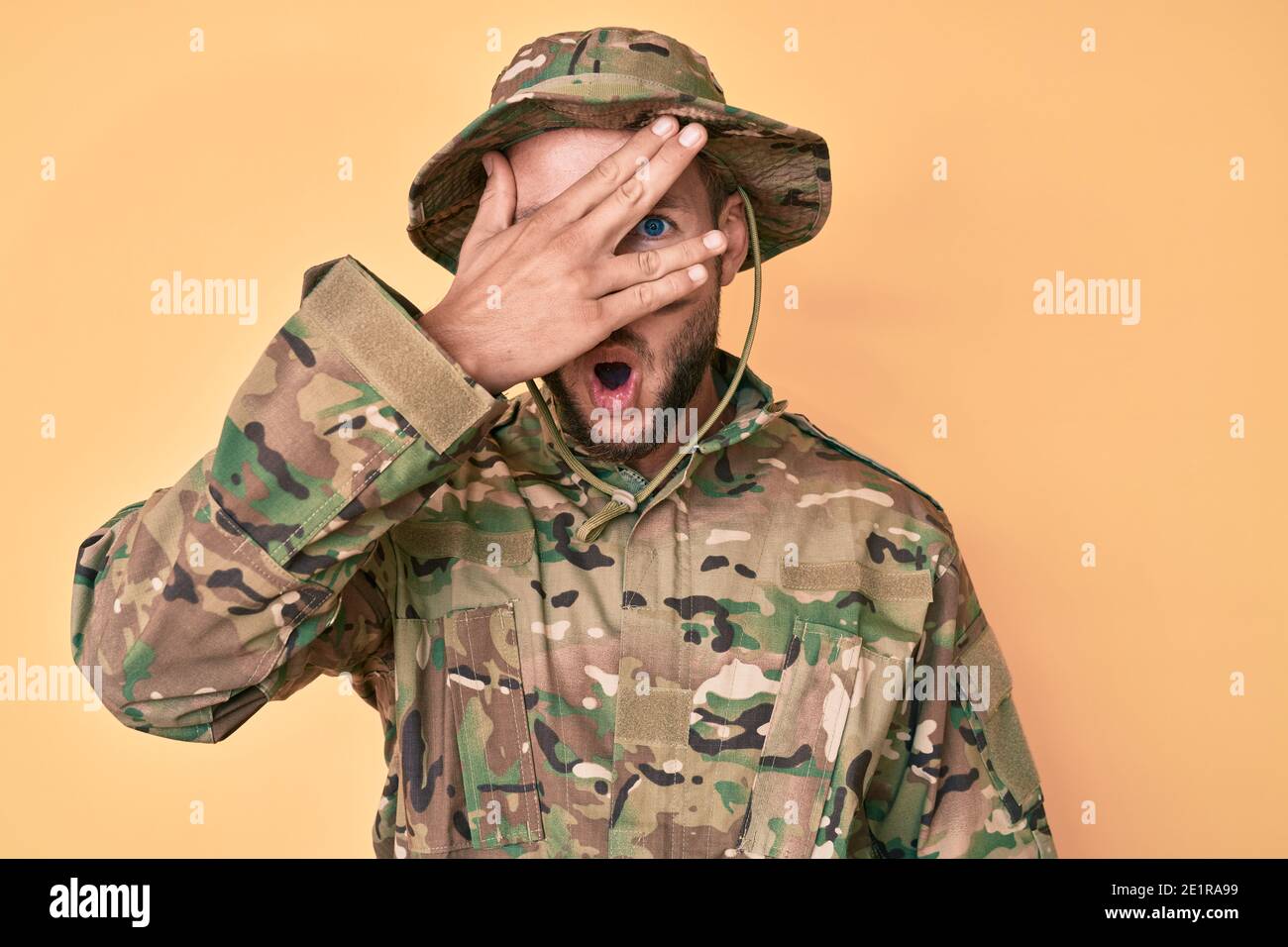 Young caucasian man wearing camouflage army uniform peeking in shock ...