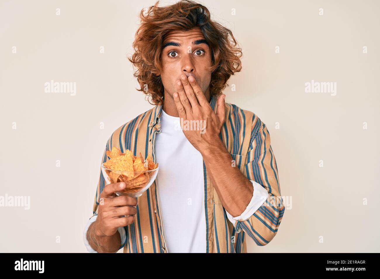 Young hispanic man holding nachos potato chips covering mouth with hand ...
