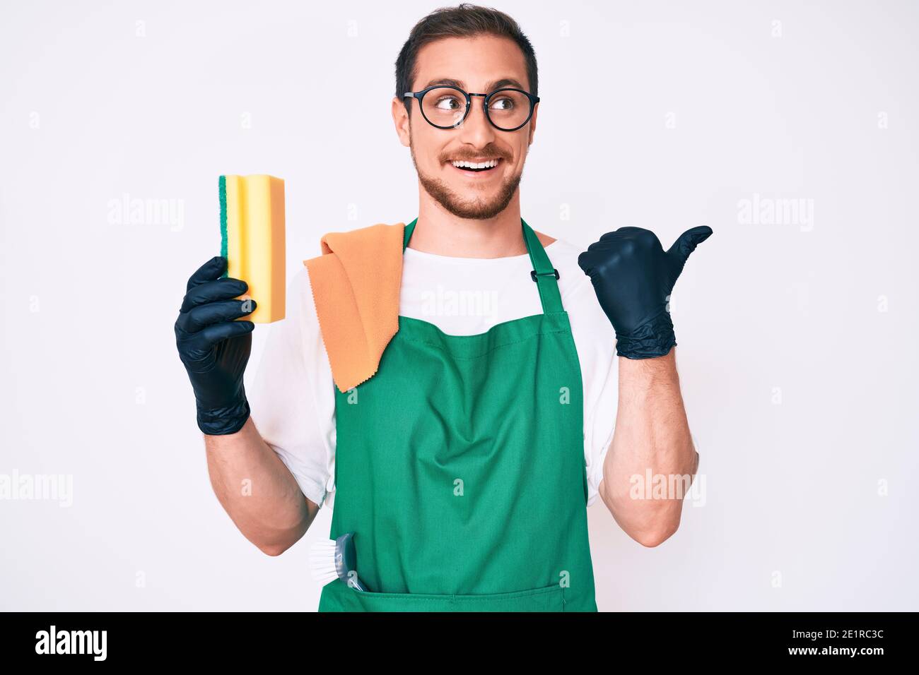 Young handsome man wearing apron holding scourer pointing thumb up to ...