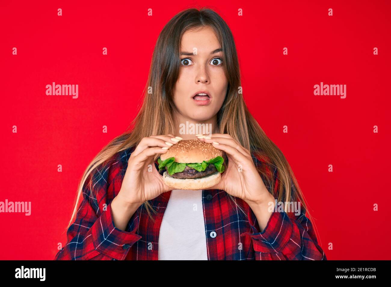 Beautiful caucasian woman eating a tasty classic burger in shock face ...