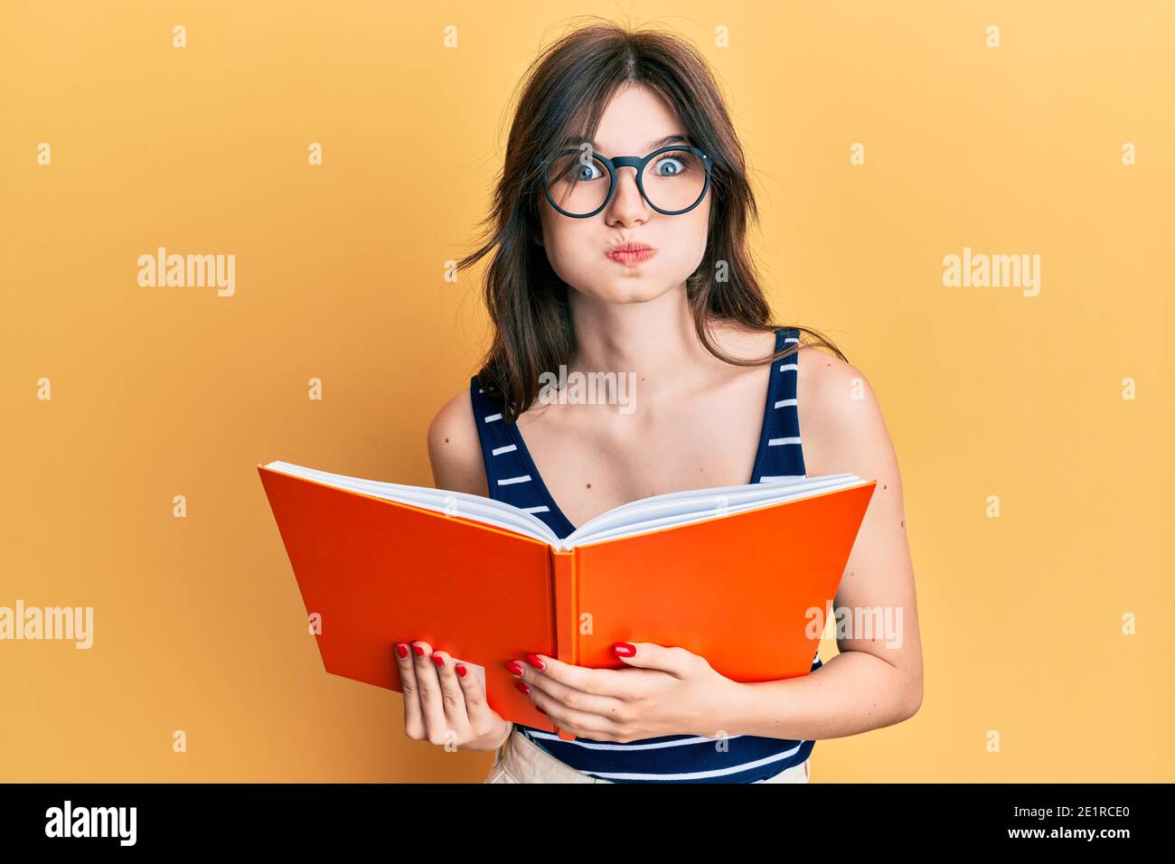 Young beautiful caucasian girl reading a book wearing glasses puffing cheeks with funny face ...