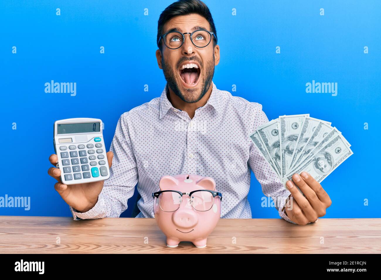 Handsome hispanic man holding dollars calculating savings angry and mad ...