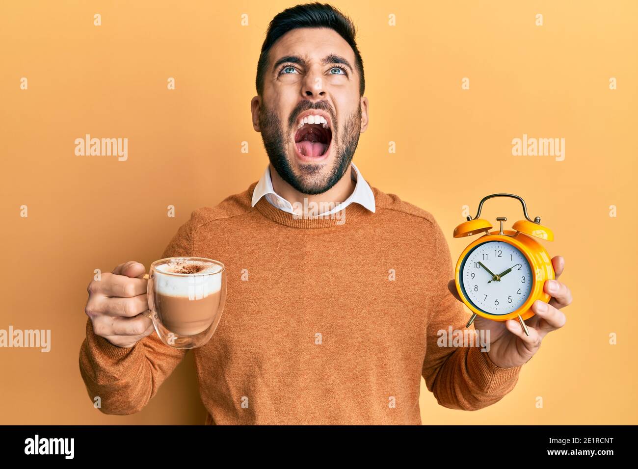 Young hispanic man holding coffee and alarm clock angry and mad ...