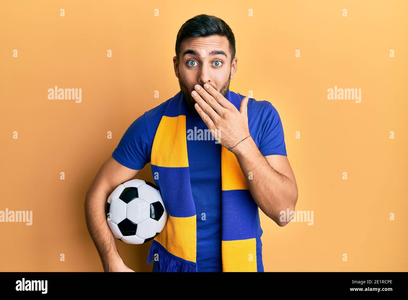 Young hispanic man football hooligan soccer ball covering mouth with ...