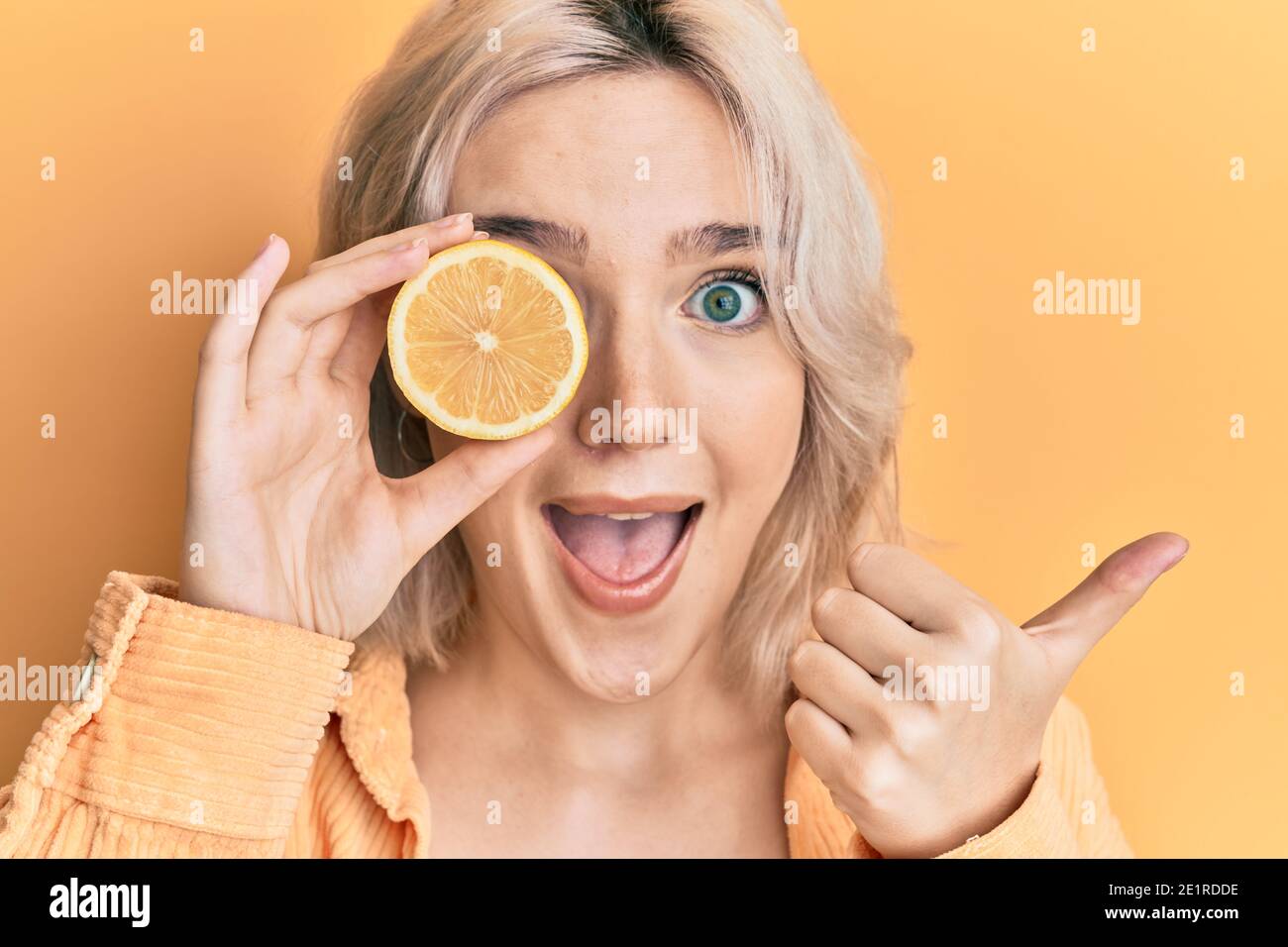 Young blonde girl holding lemon over eye pointing thumb up to the side ...